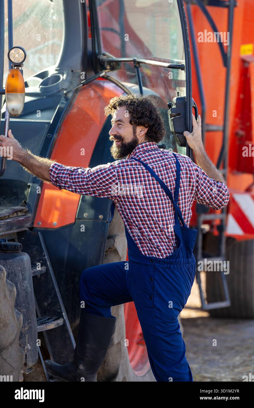 Farmer in overalls and with rubber boots climbing on the tractor's ...