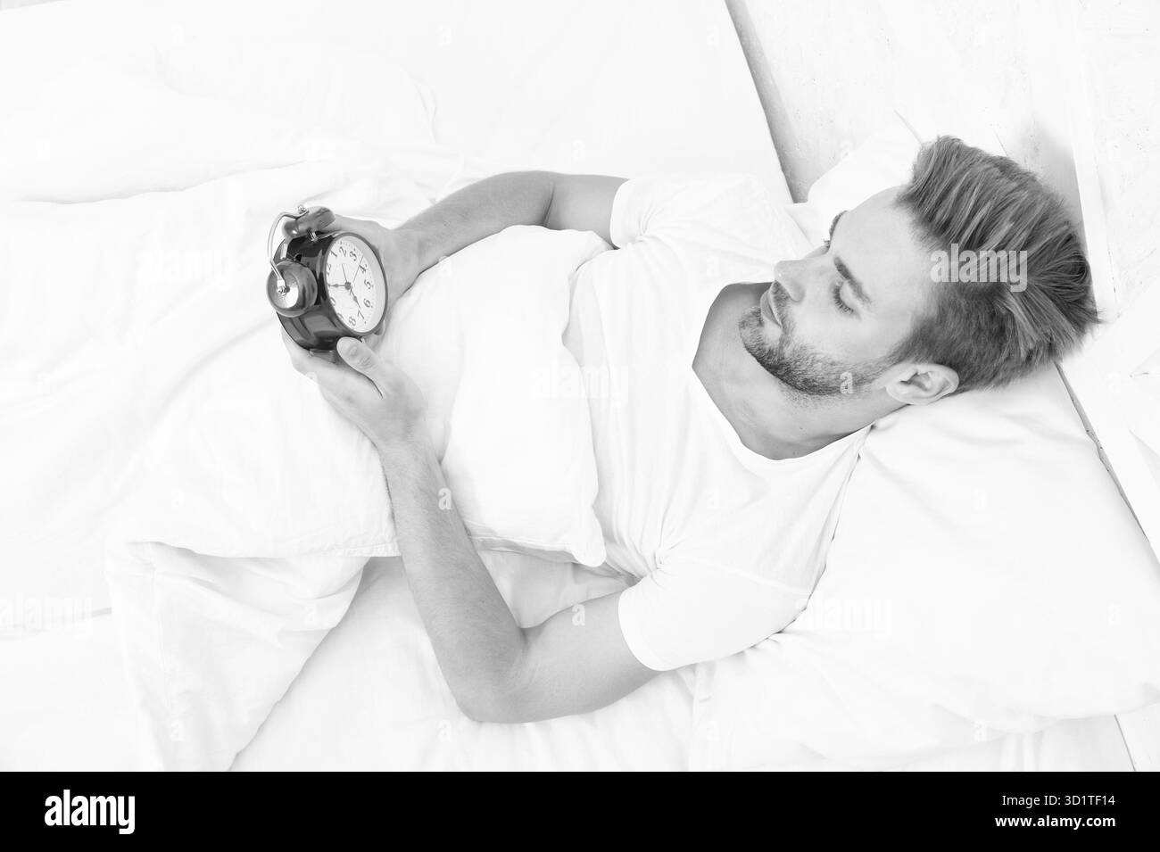 Sleepy man looking at alarm clock in hand while lying in bed. Morning ...