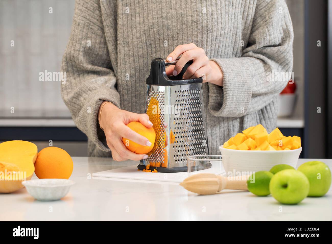 Caucasian adult grating fresh orange in kitchen with fruit selection ...