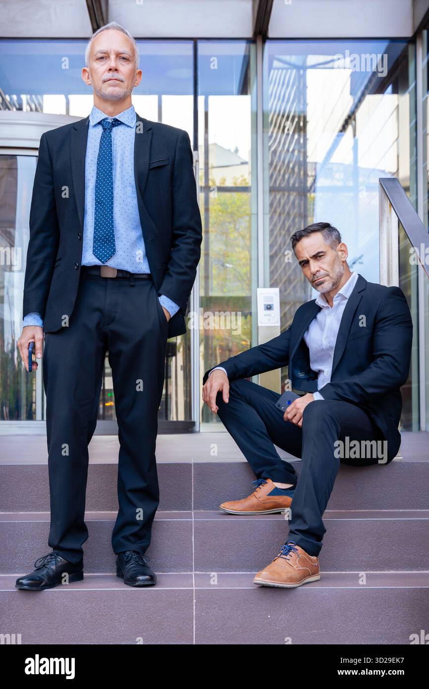 Two businessmen in suits posing outside modern office building, serious ...