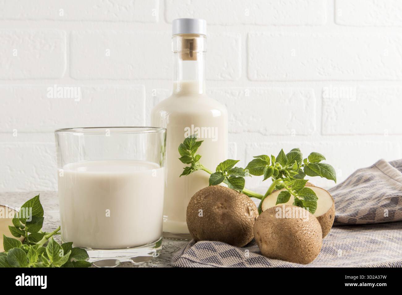 A glass and bottle of vegetable potato milk against a white brick wall ...