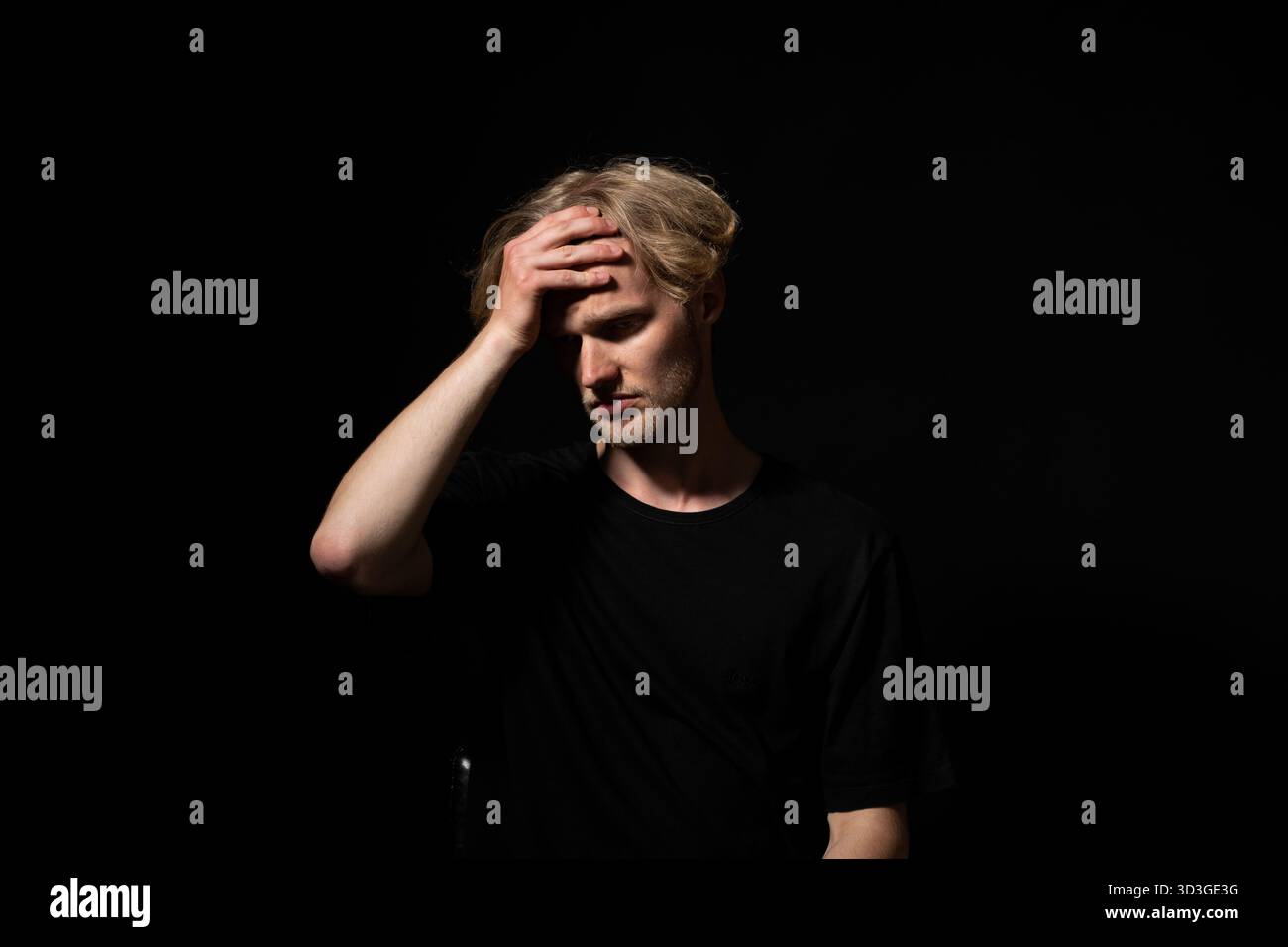 Sad man in black shirt holding head and closing eyes in dark studio ...