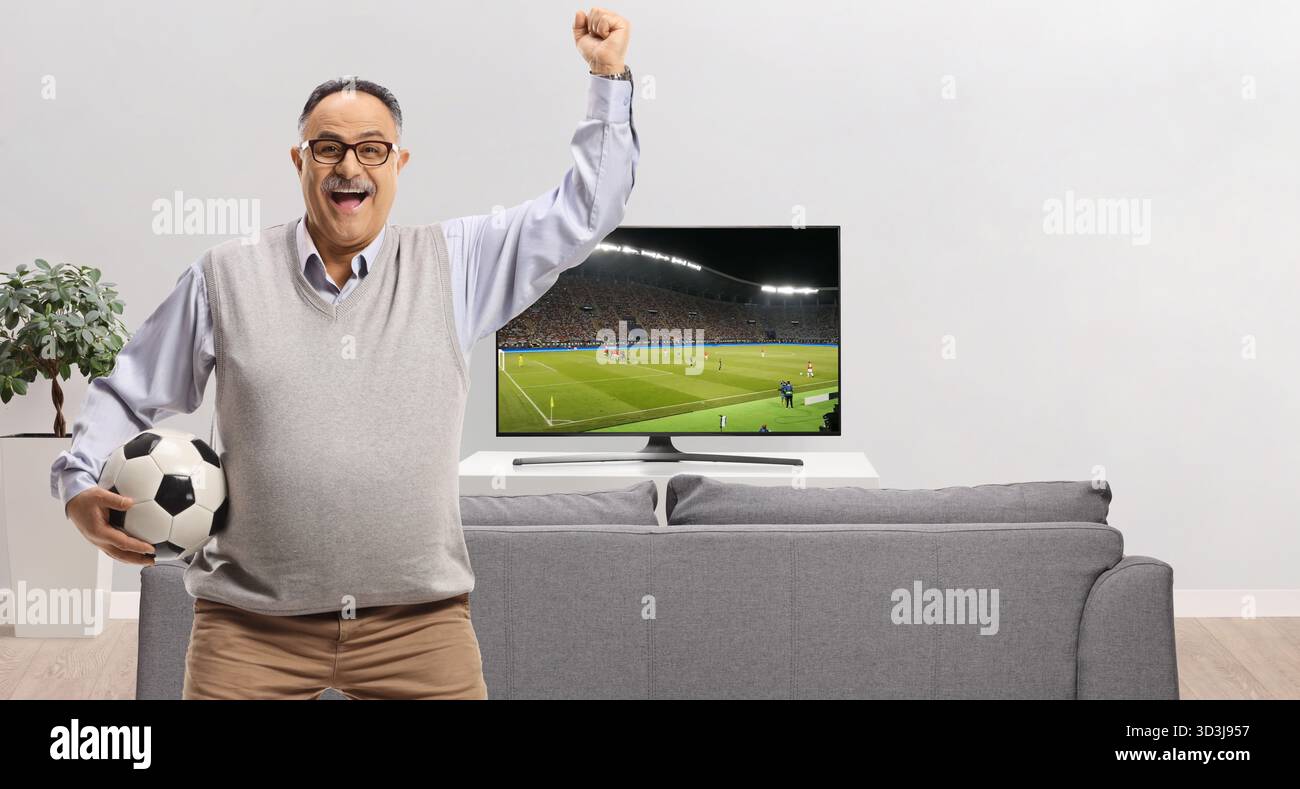 Elderly football fan cheering with a ball in his hands in a room with ...
