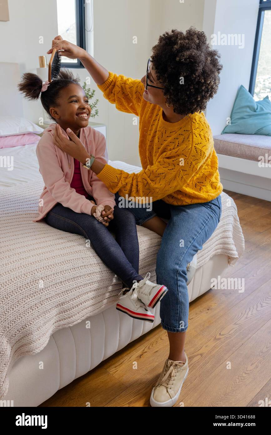 Brushing Diverse mother and daughter sitting on edge of bed in modern ...