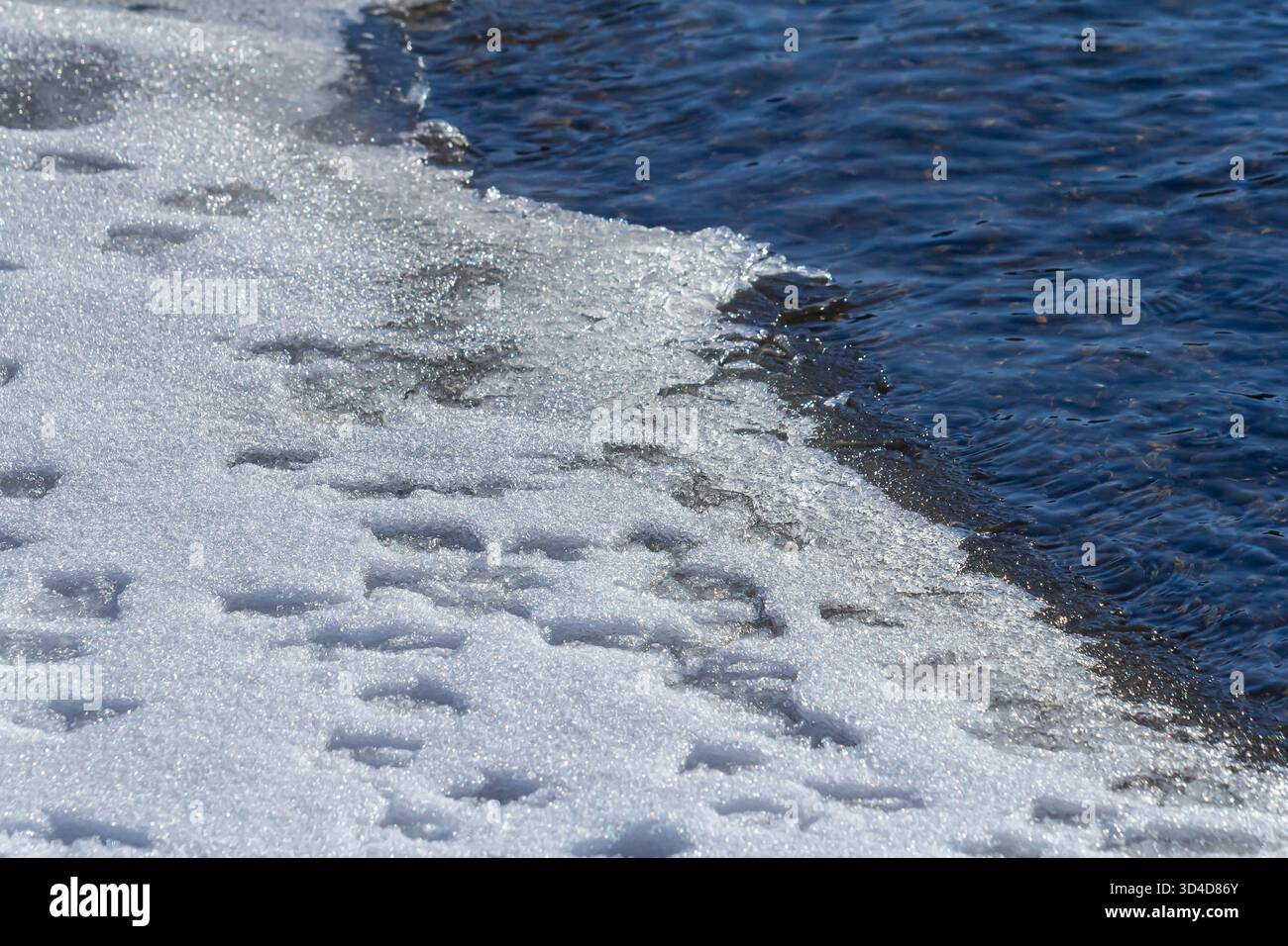 Snow and ice partially cover the edge of a calm river with clear blue ...