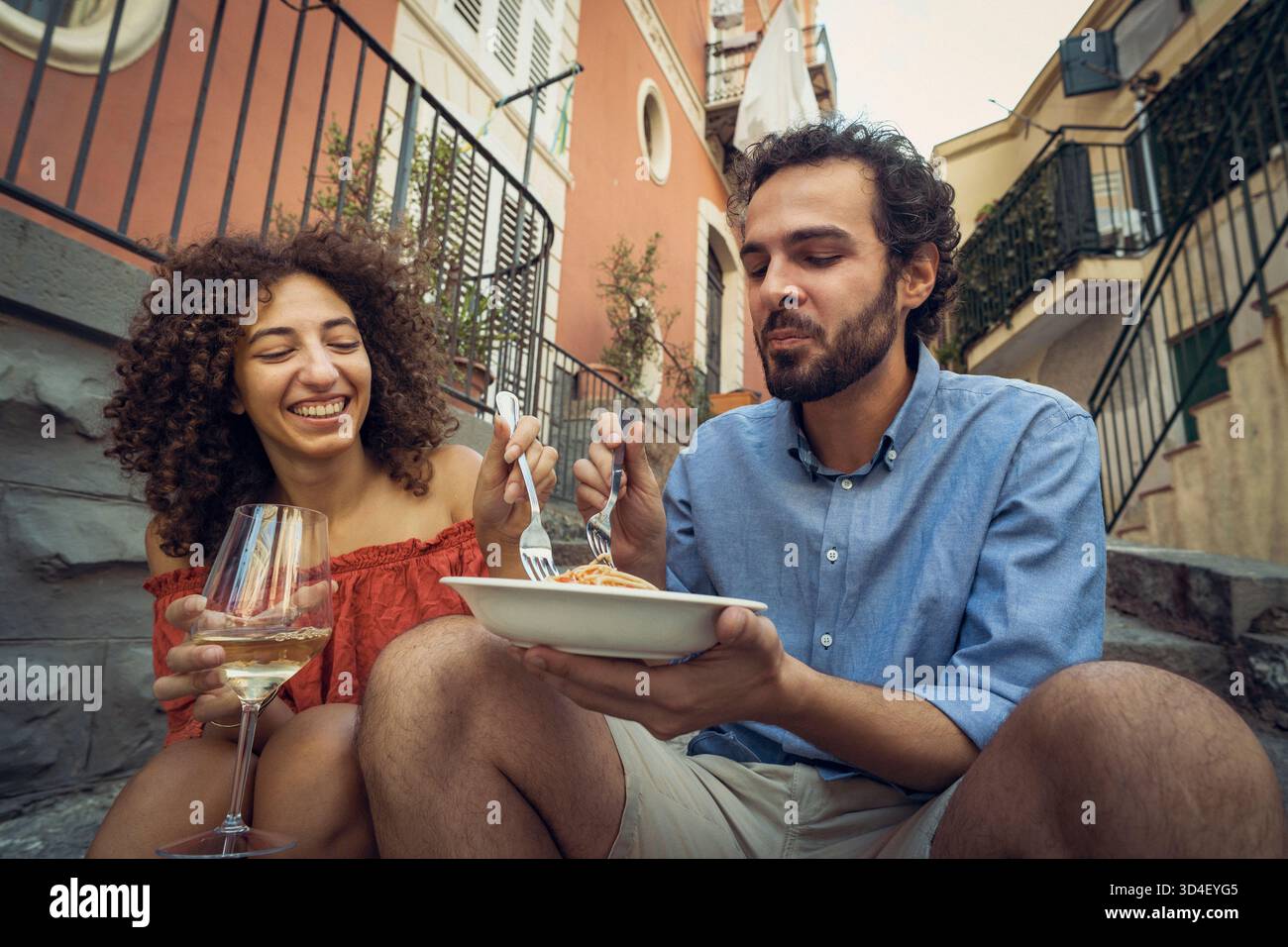 Young couple eating pasta and drinking wine while sitting on the steps ...