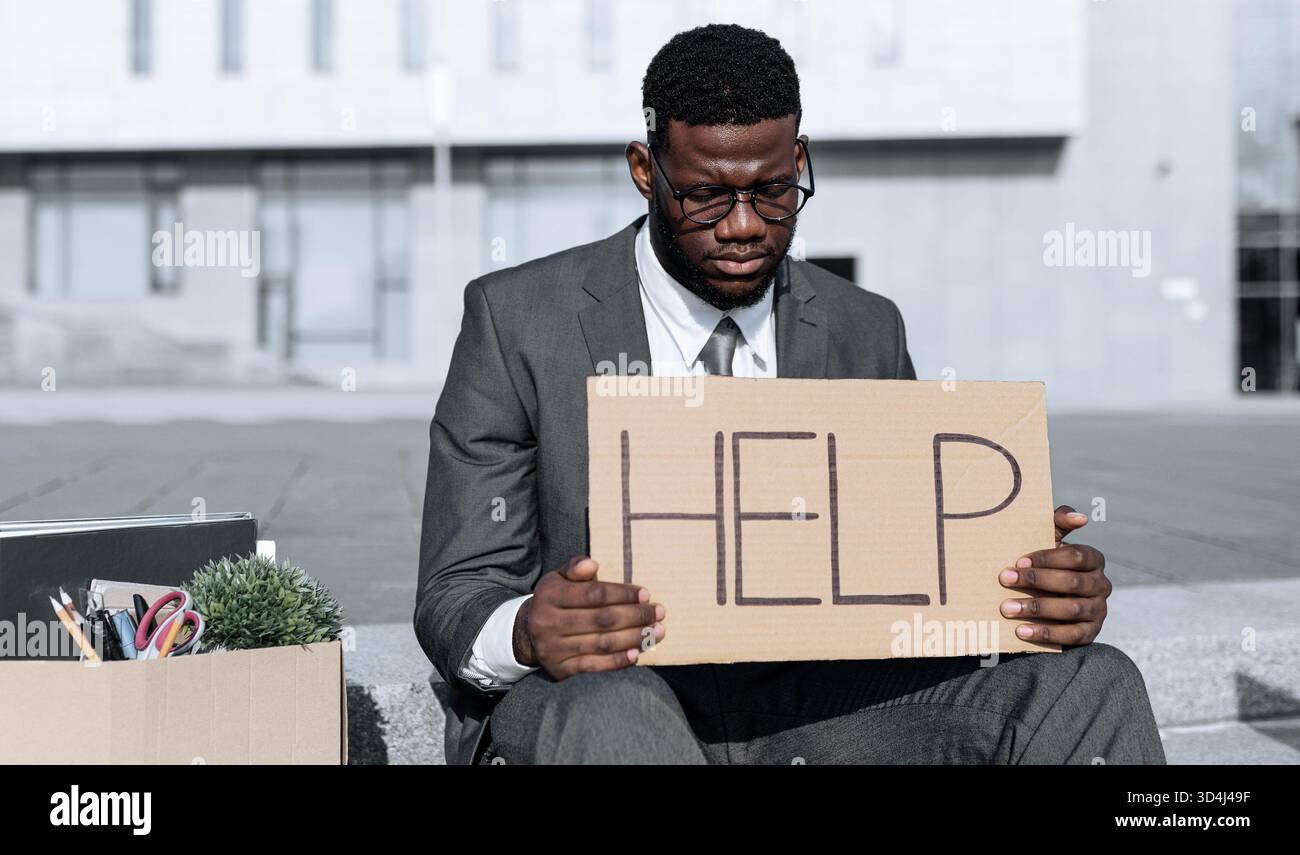 Man in Suit Holding Help Sign Sits on Sidewalk With Supplies Beside Him ...