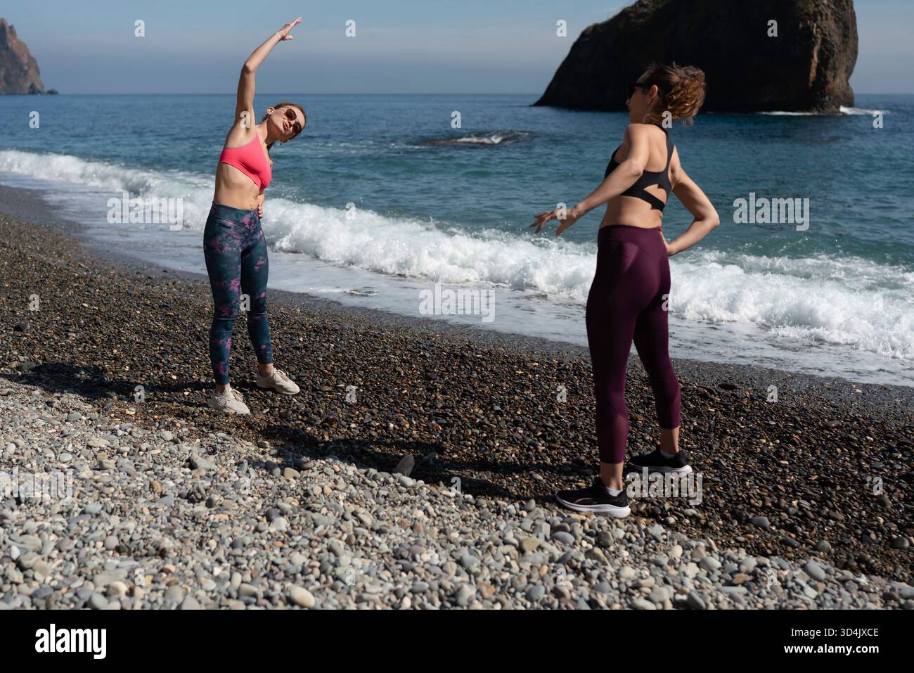Two women stretch on a rocky beach near the ocean. One in a pink top ...