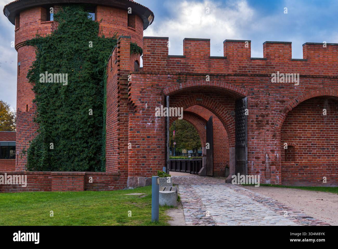 Red brick gatehouse with ivy clad round tower stands by cobbled ...
