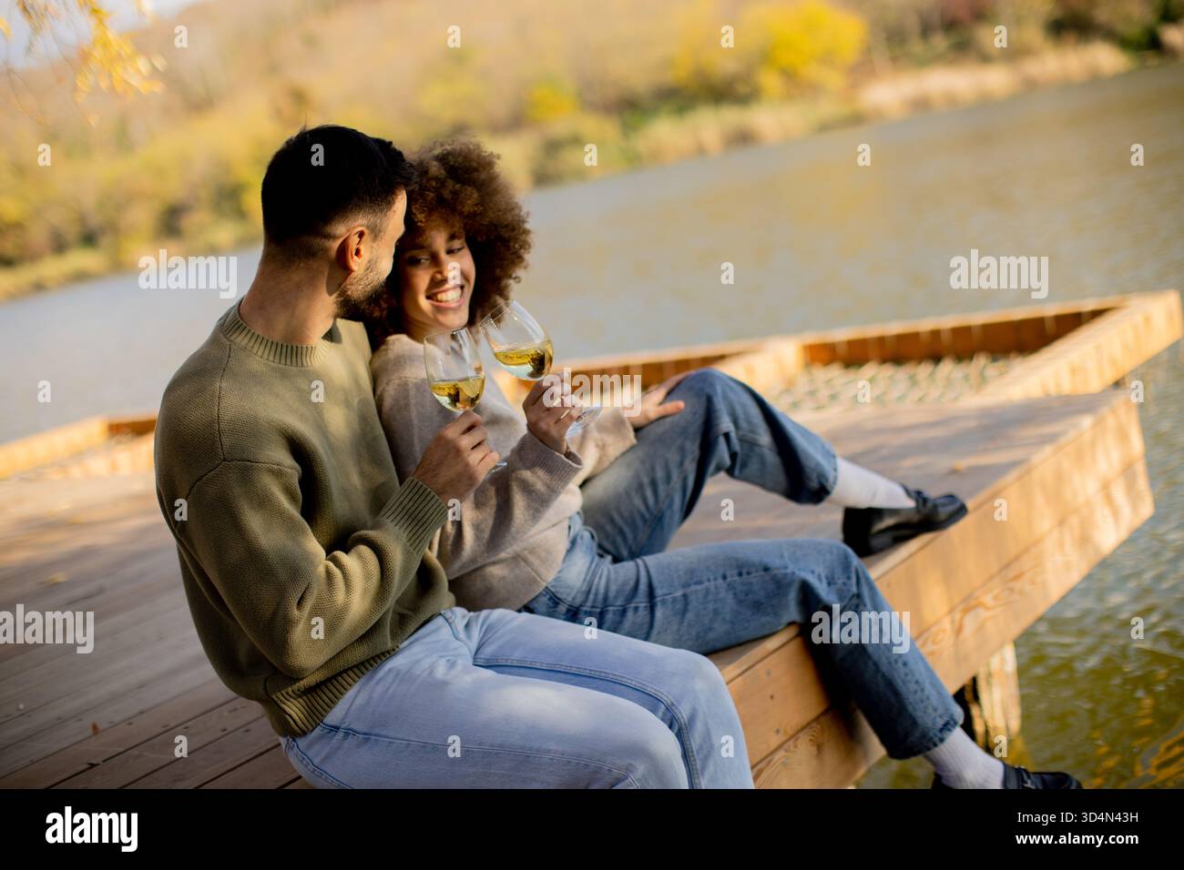 Two people sit together on a wooden dock, smiling and holding glasses ...