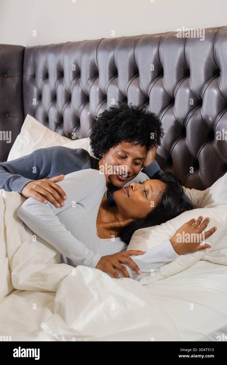Snuggling Diverse couple lying on bed in softly lit bedroom, with white ...