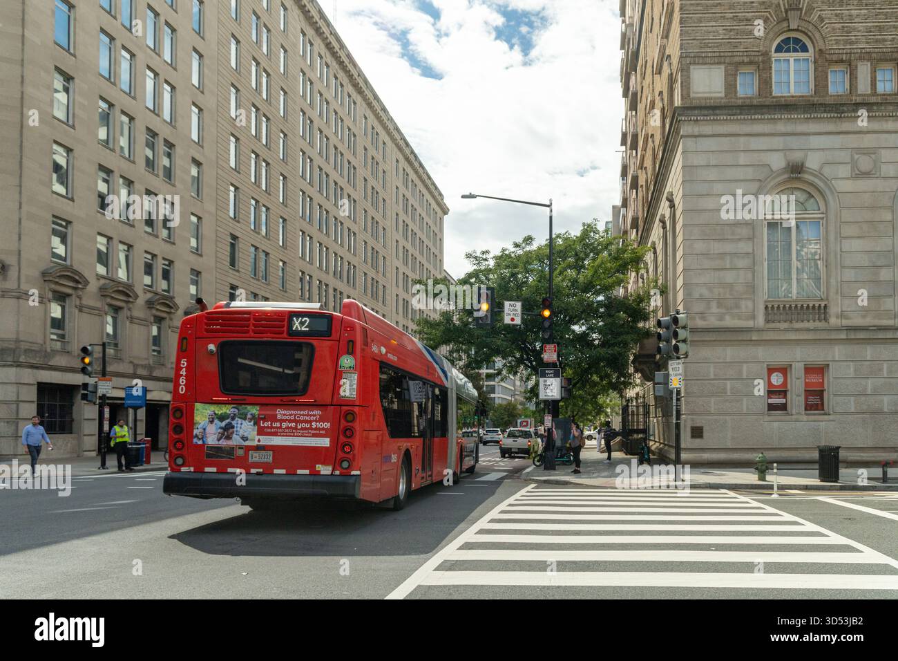 A red bus is driving down a city street. The bus is surrounded by tall ...