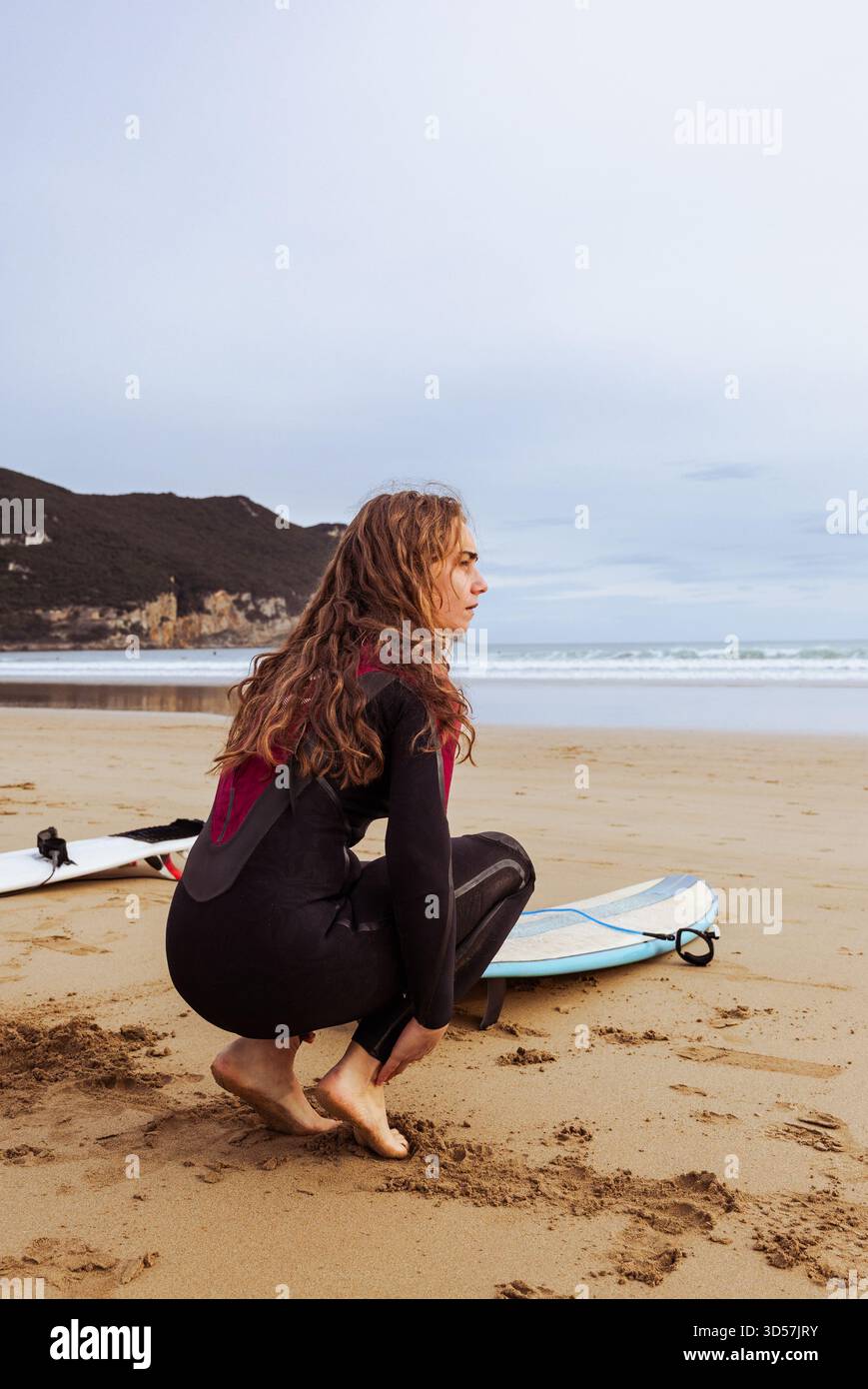 Female surfer squatting on the sand next to her board watching the ...