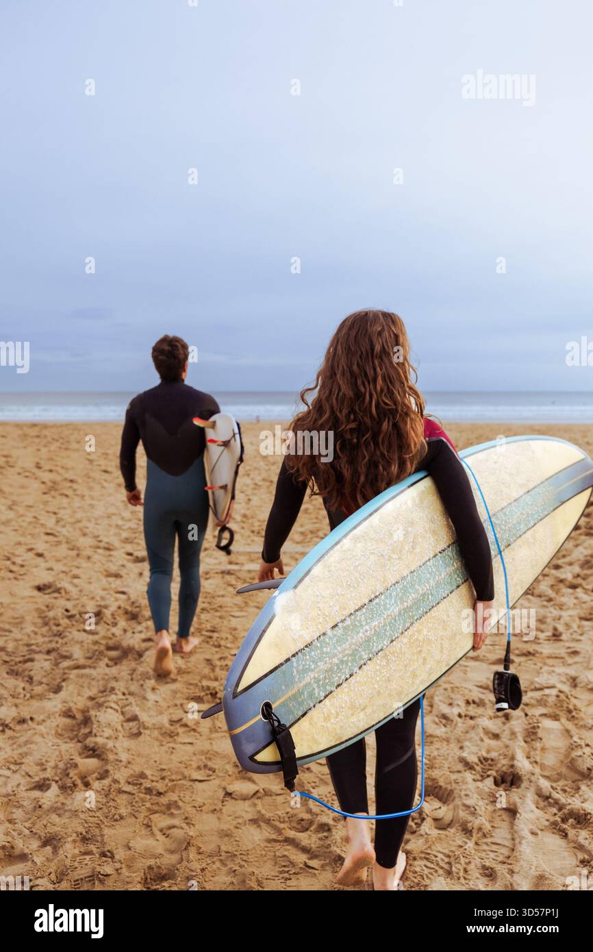Two friends in wetsuits walk barefoot on sand carrying surfboards ...