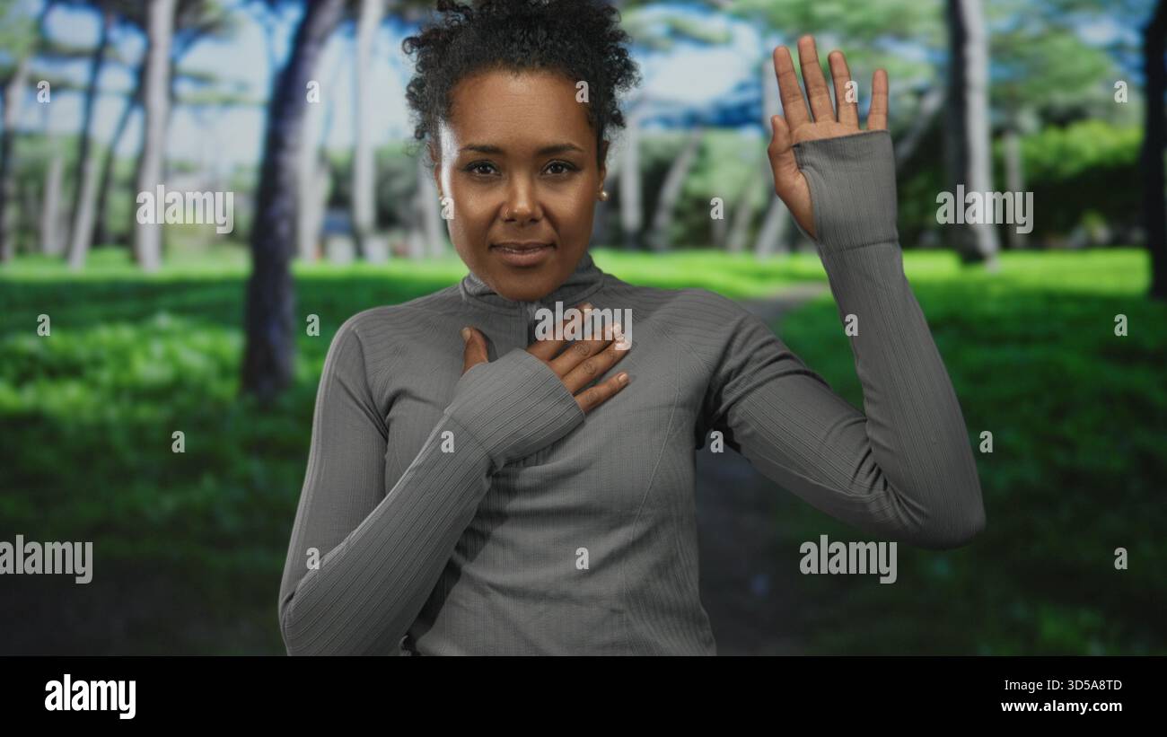 Woman stands amid green trees in a forest raising her hand in greeting ...