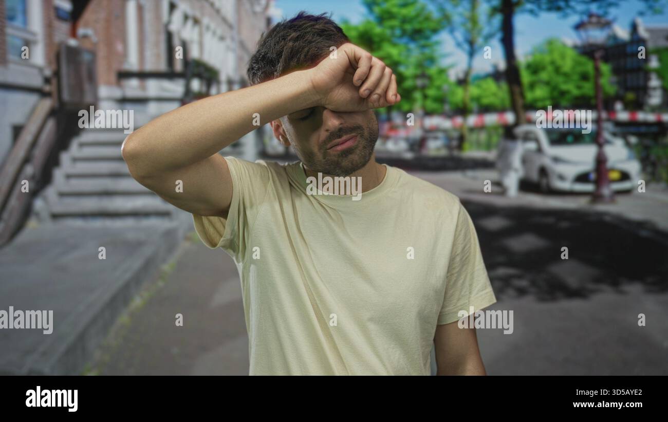 Man wipes sweaty forehead on sunlit city street with brick building ...
