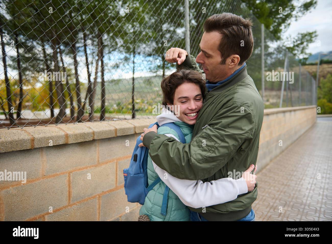 A father warmly hugs his teenage son in an outdoor setting by a fence ...