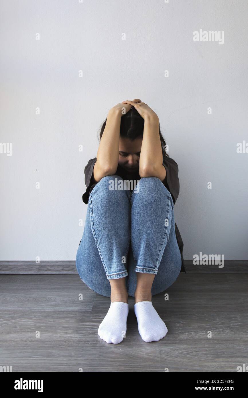 A woman sitting on the floor against a wall, hugging her knees and ...