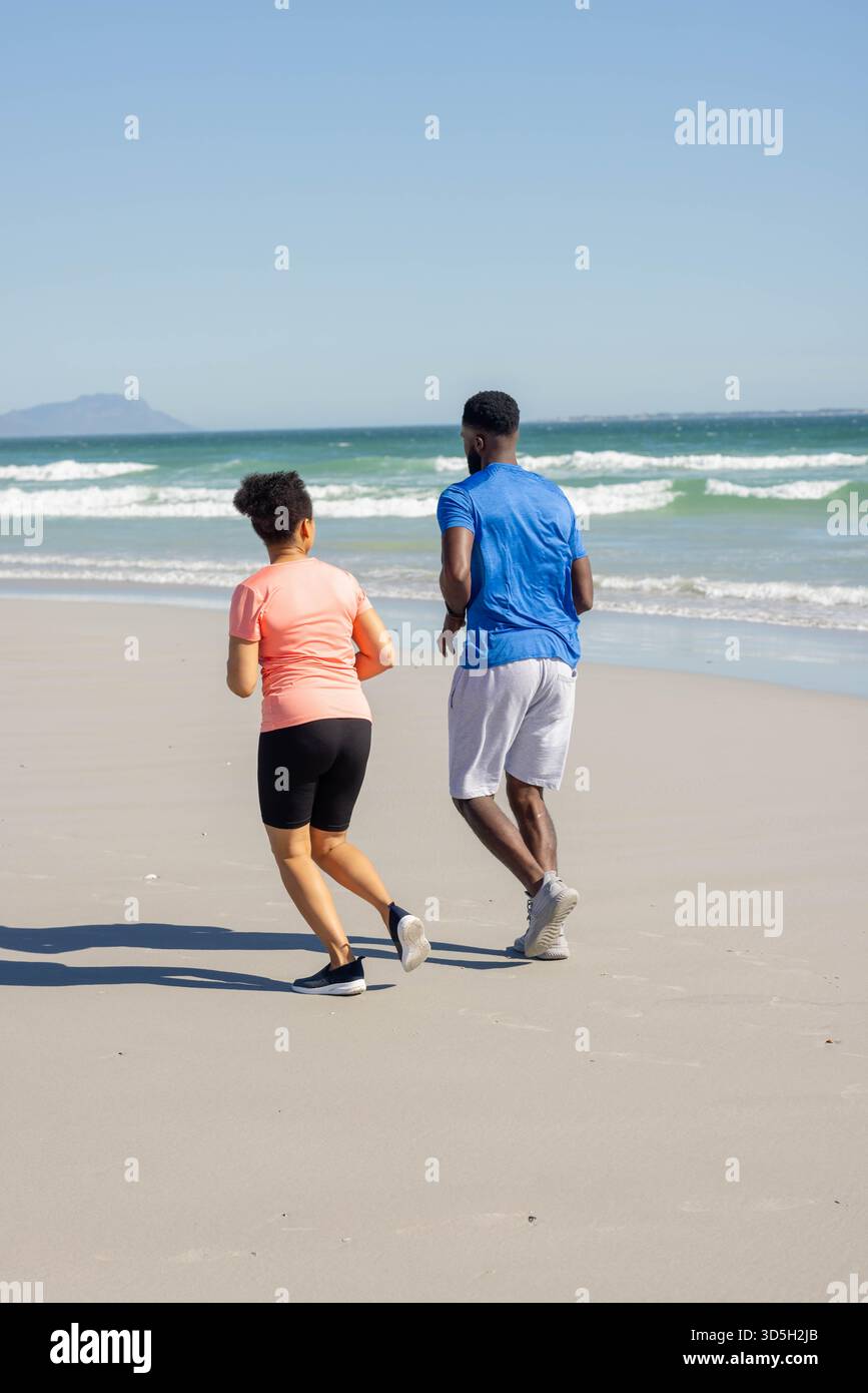 African American couple jogging on beach, enjoying sunny day by ocean ...