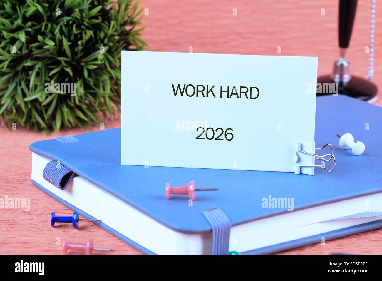 A blue notebook lies on a wooden table next to a small plant and a pen ...
