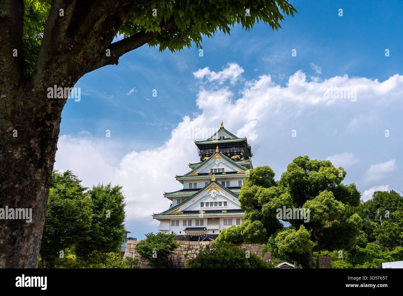 The majestic main tower of Osaka Castle rises above green trees under a ...