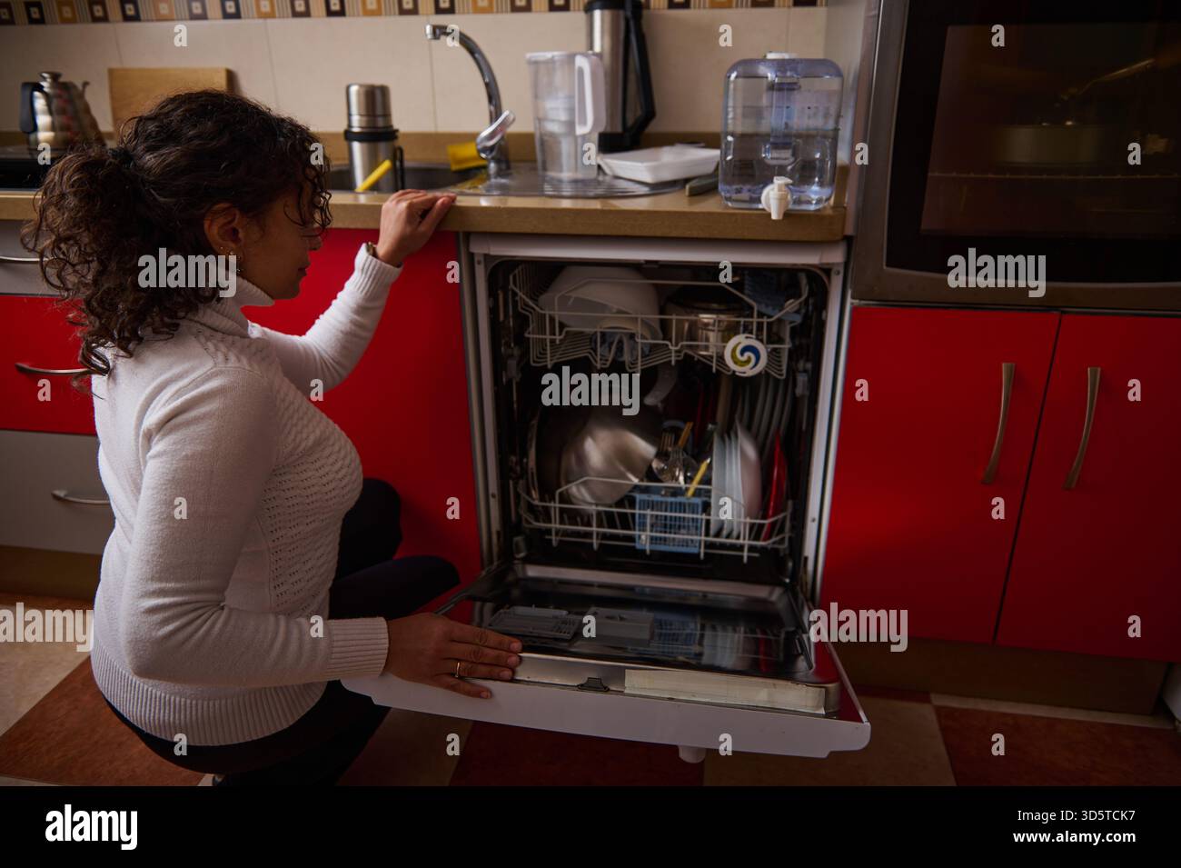A woman kneels beside an open dishwasher in a bright red kitchen ...