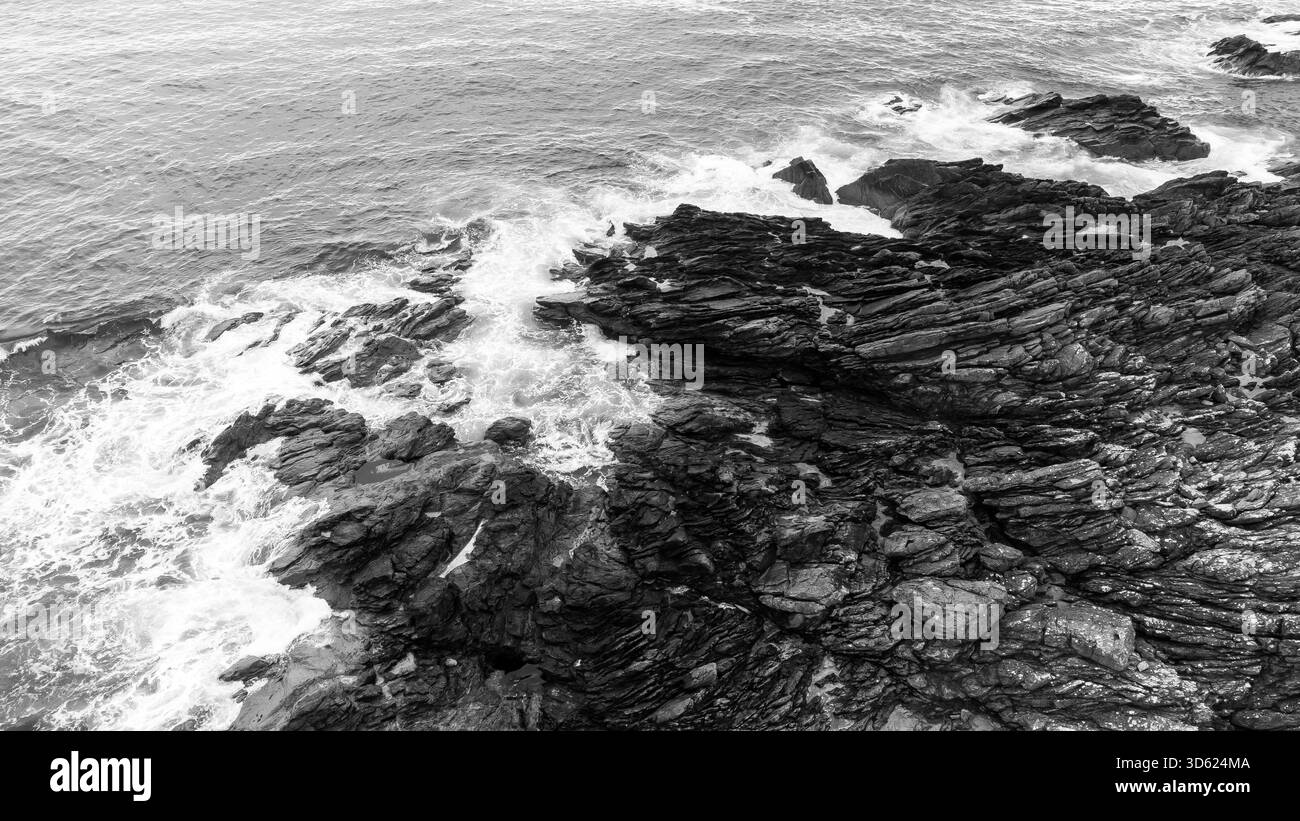 Aerial monotone view of rugged coastal rocks with waves breaking below ...