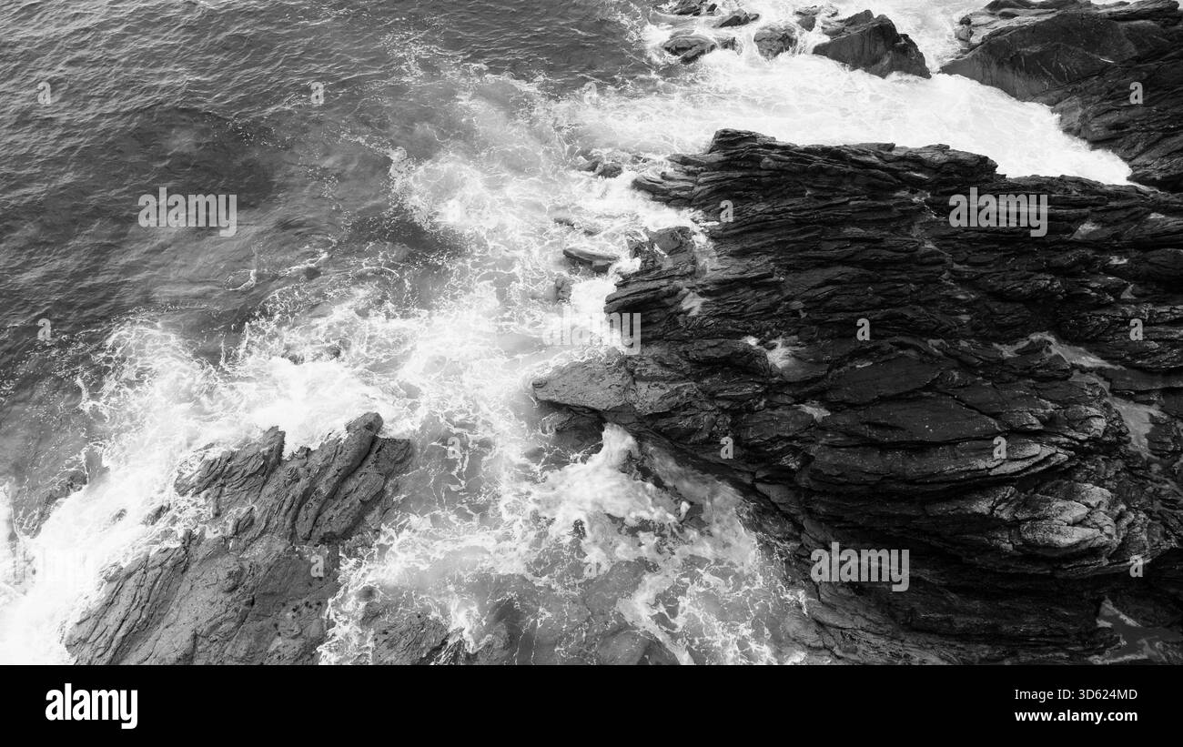 Aerial monotone view of rugged coastal rocks with waves breaking below ...