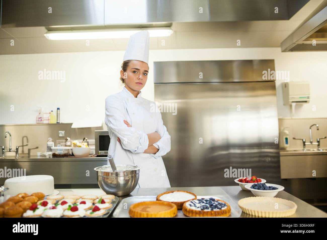 Woman chef wearing white coat and tall toque, standing at prep table ...
