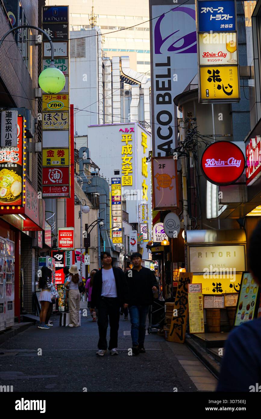 Pedestrians walk through a bustling side street in Shibuya filled with ...