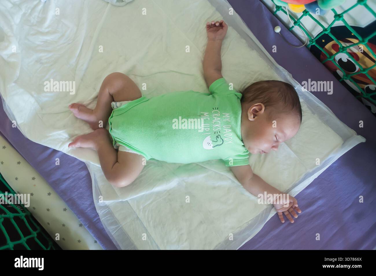 A sleeping baby in a green onesie rests on a soft surface in a well-lit ...
