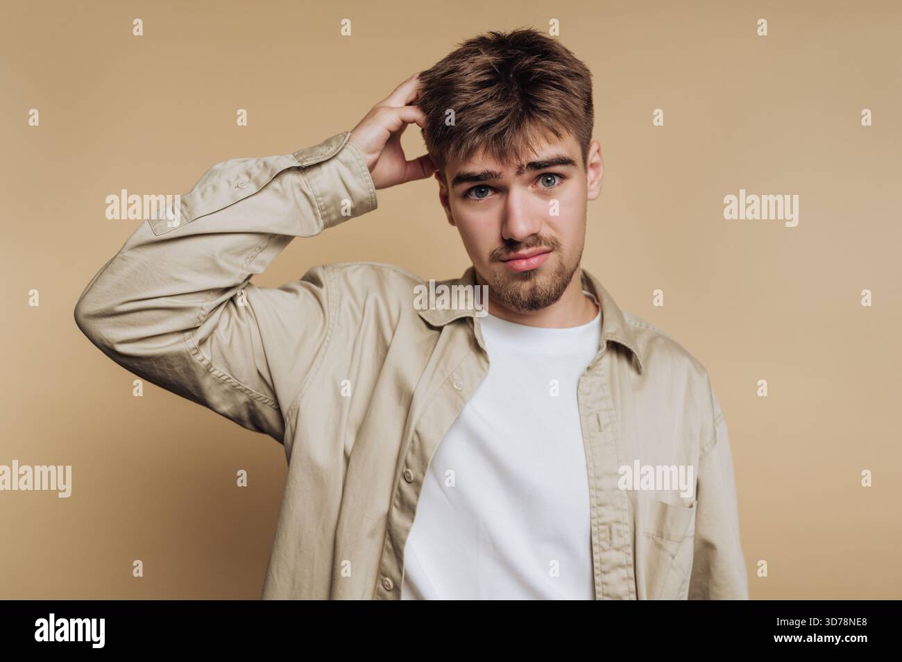 A young man stands against a plain beige backdrop, looking puzzled and ...
