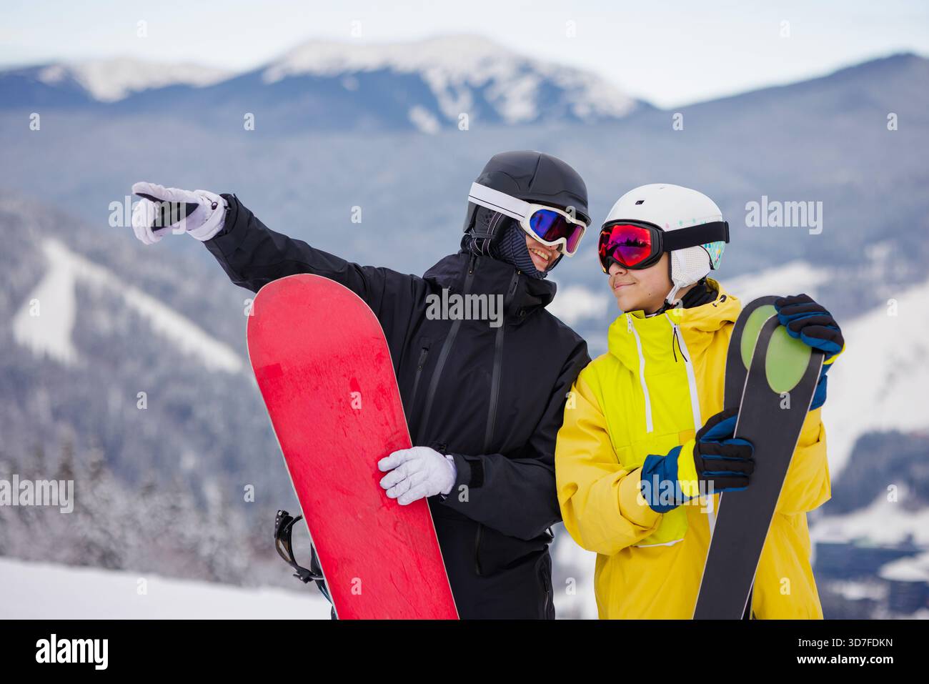Smiling teenage snowboarder pointing out route to skier friend on snowy ...