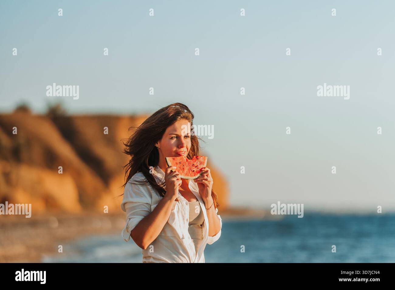 A beautiful young woman in a white shirt walks on the beach by the sea and eats watermelon at ...