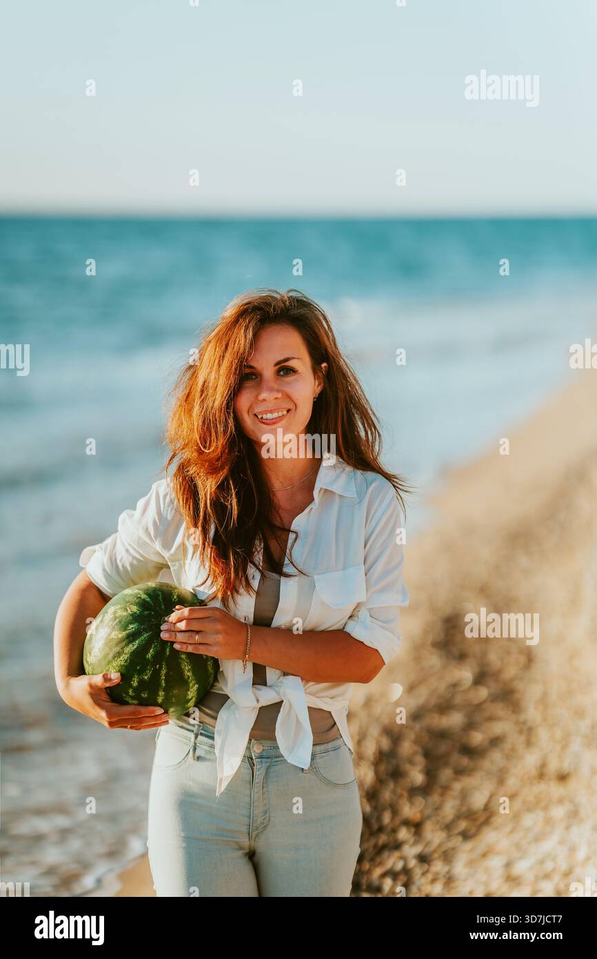 A beautiful young woman in a white shirt walks on the beach by the sea and eats watermelon at ...