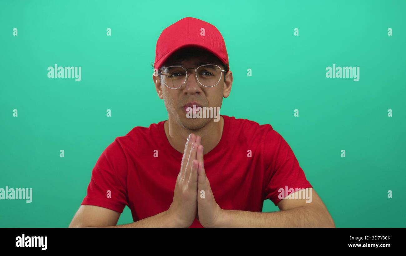 Young hispanic man in red shirt and cap holds hands pressed in prayer ...