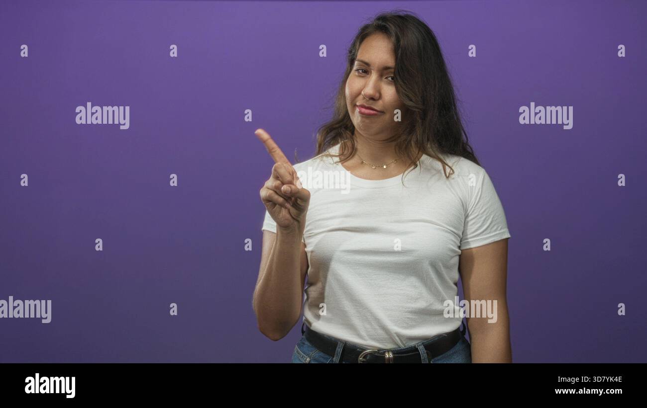 Woman pointing index finger to side in purple studio wearing white tee ...