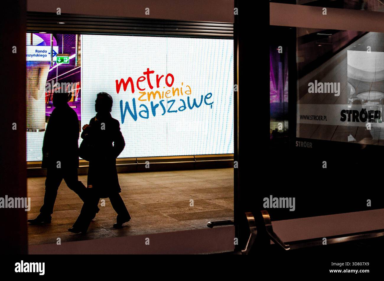 Silhouetted pedestrians passing a bright display reading "Metro zmienia ...