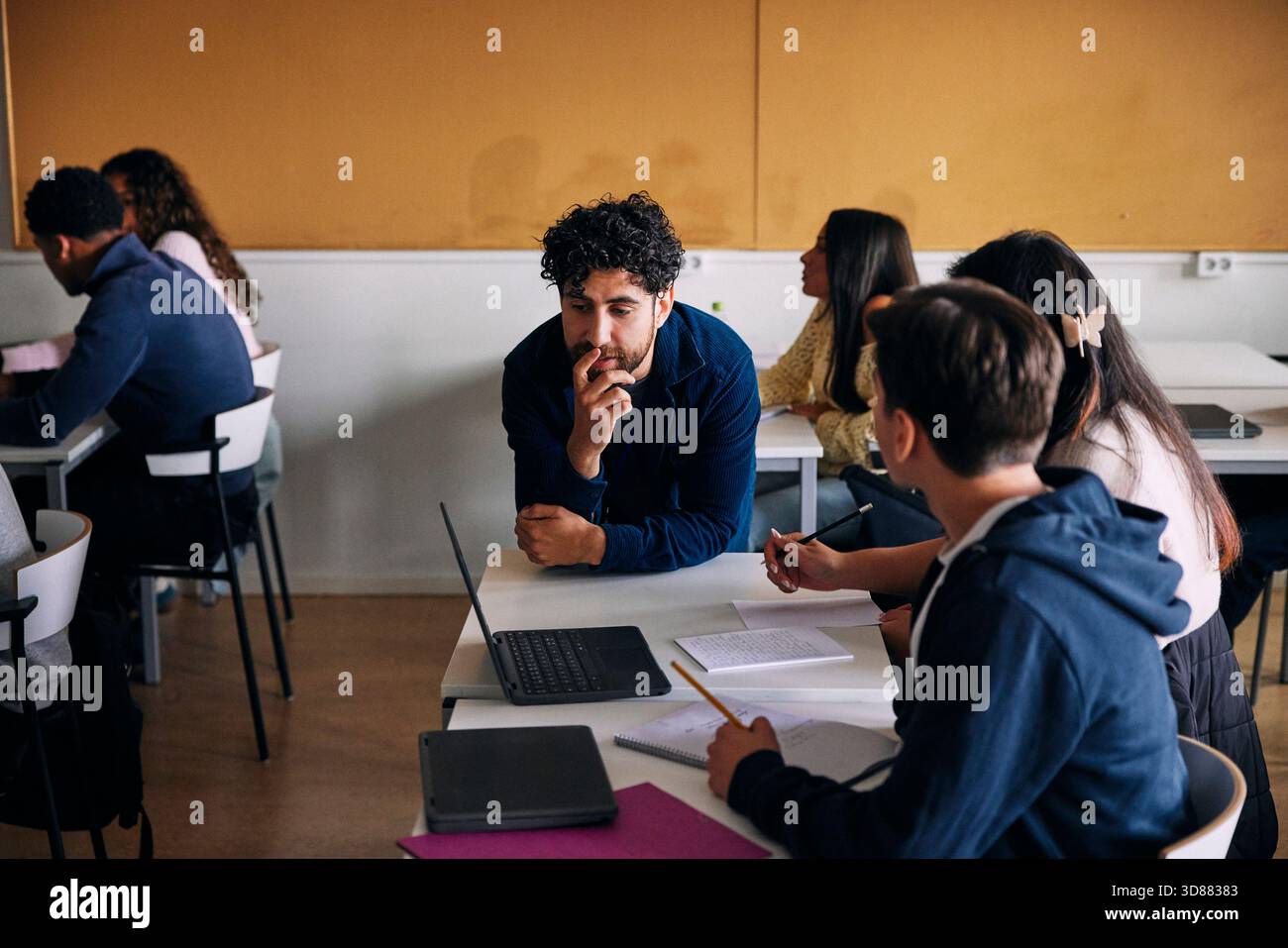 Male teacher assisting high school students while leaning on desk in ...
