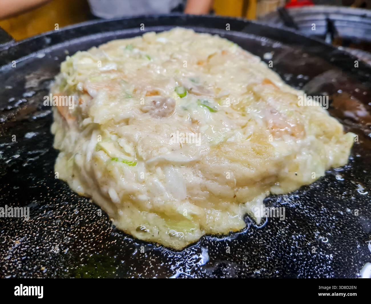 A thick okonomiyaki patty cooks on a sizzling griddle, with bubbling ...