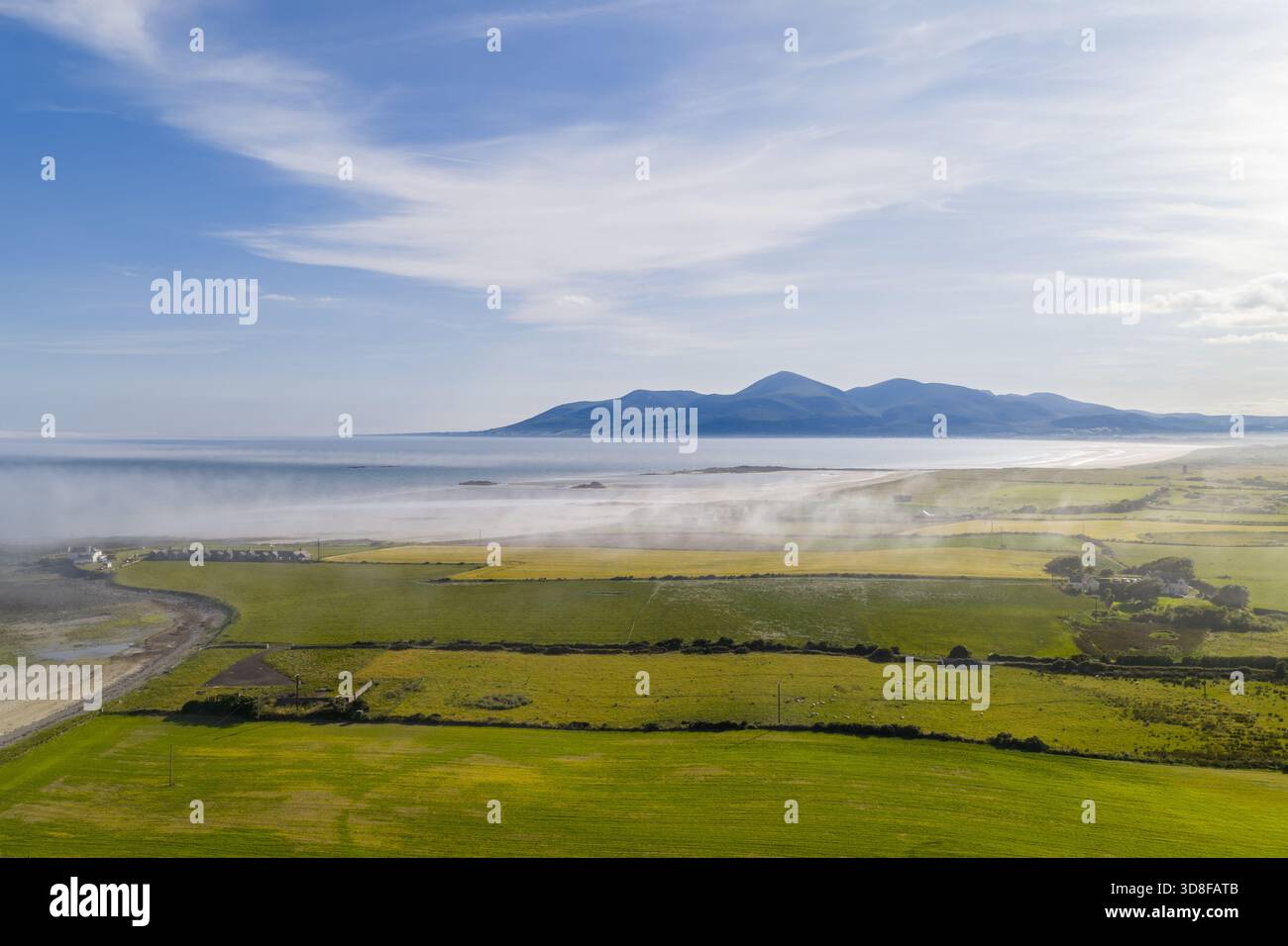 A serene coastal landscape shows vibrant green fields, a misty ...