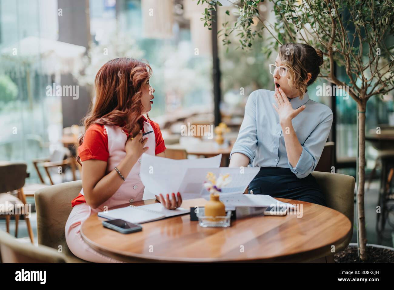 Two women discuss documents at a cafe, sharing ideas and surprised ...