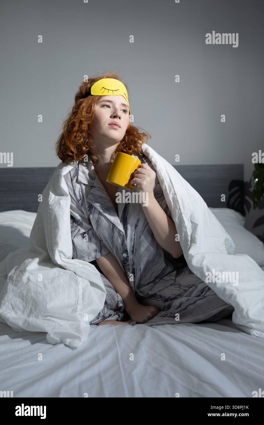 white woman on bed holding mug, wrapped in blanket with yellow eye mask ...