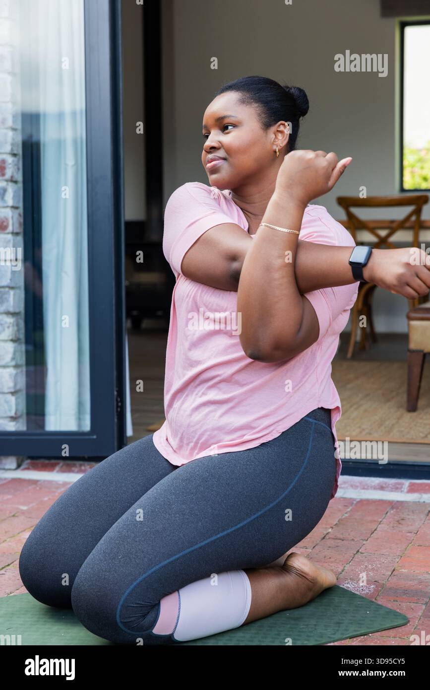 African American woman kneeling on yoga mat stretching arm across chest ...