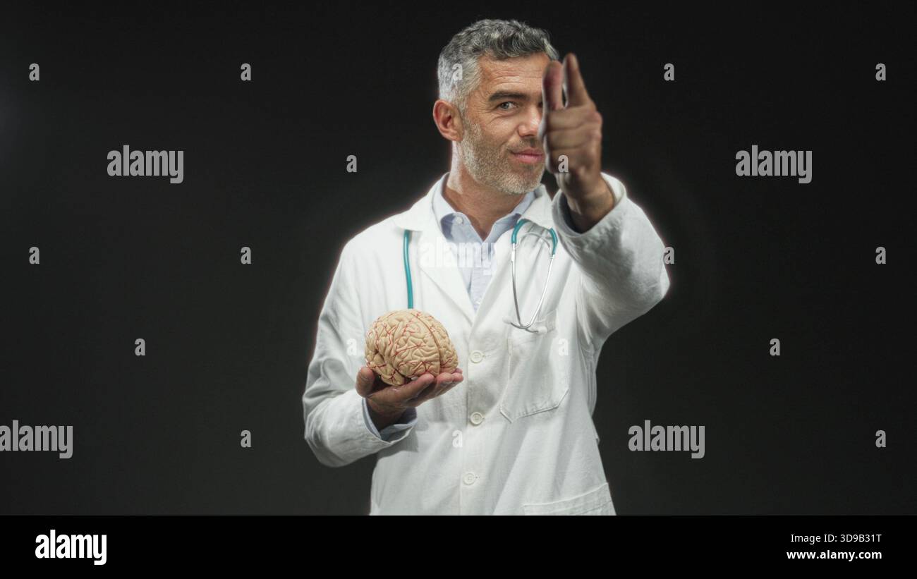 Middle aged man doctor with grey hair holds brain model in one hand and ...