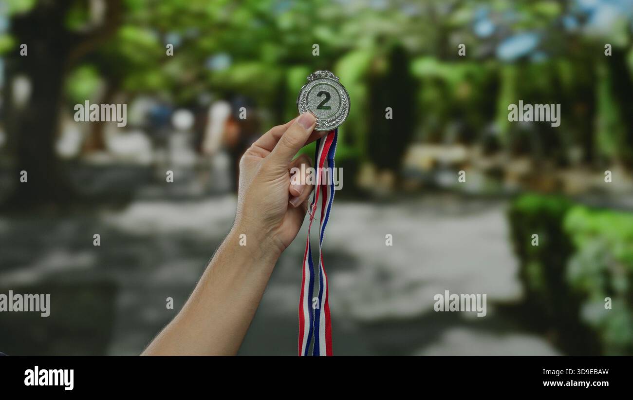 Hand holding silver medal with ribbon against park background suggests ...