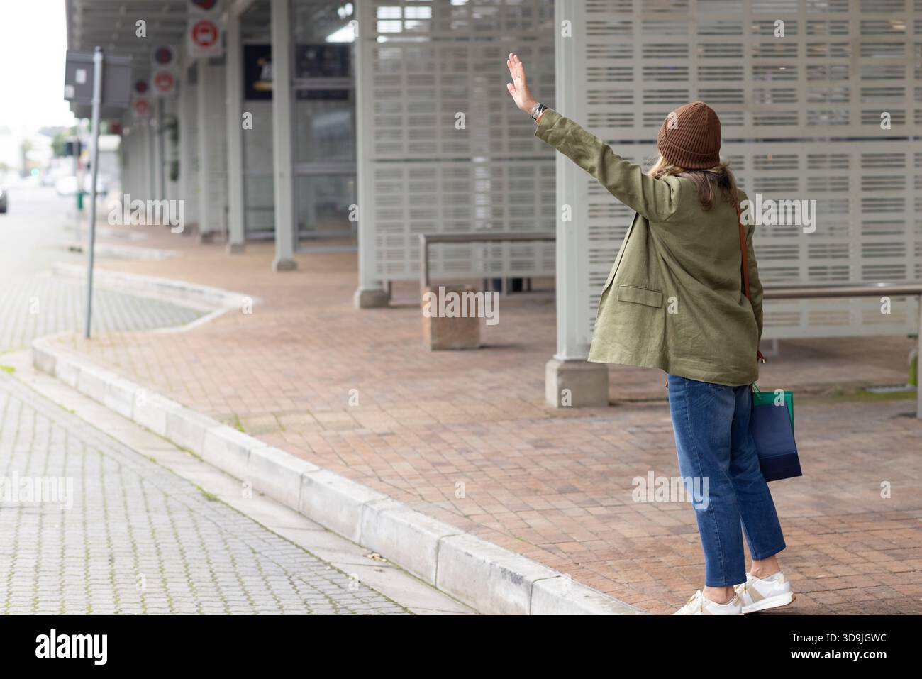 Woman raising arm and hailing bus at station platform with canvas tote ...
