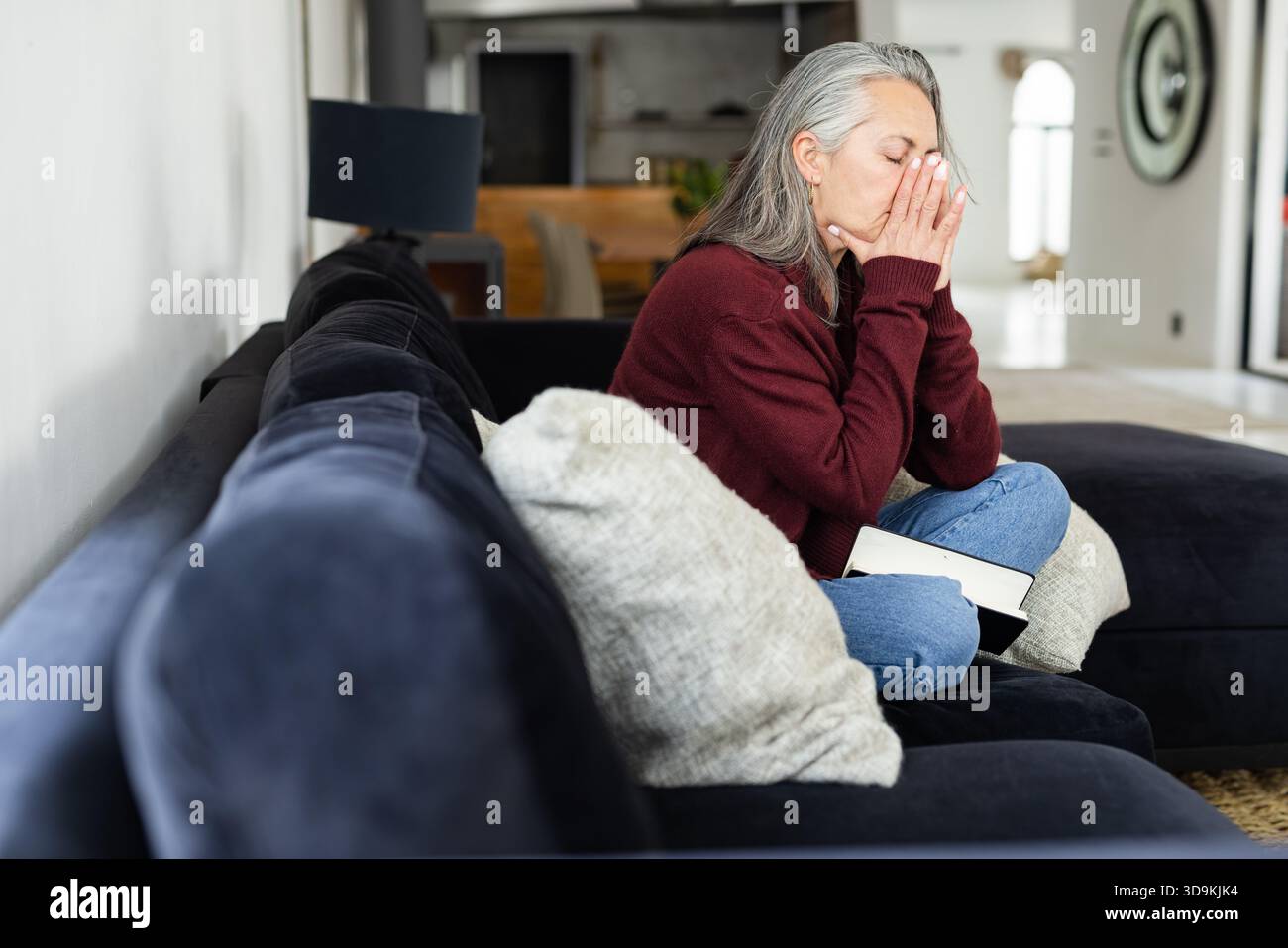 Senior woman sitting on dark-blue sofa in open-plan home pressing hands ...