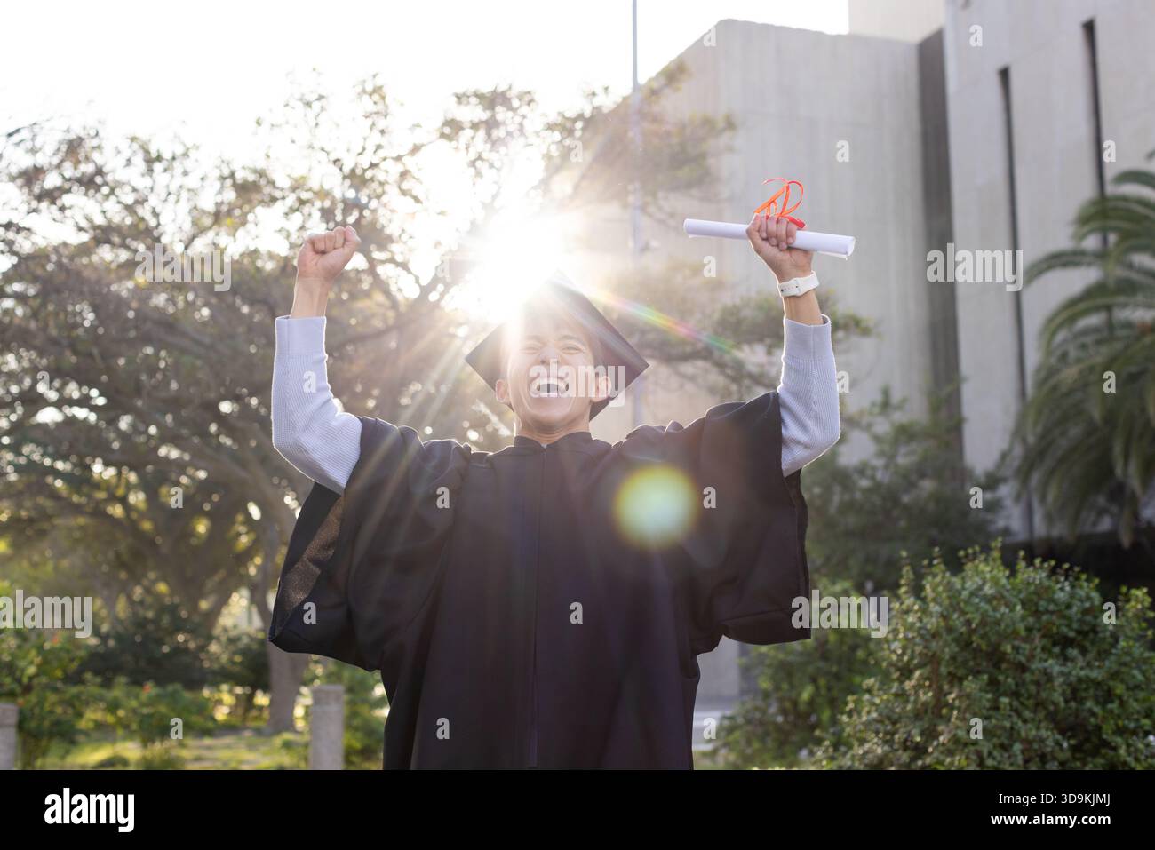 Graduate raising mortarboard and diploma scroll on landscaped campus ...