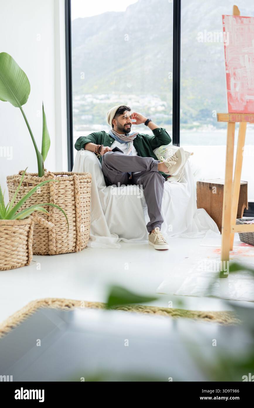 Indian man sitting on armchair holding tablet in art studio while ...